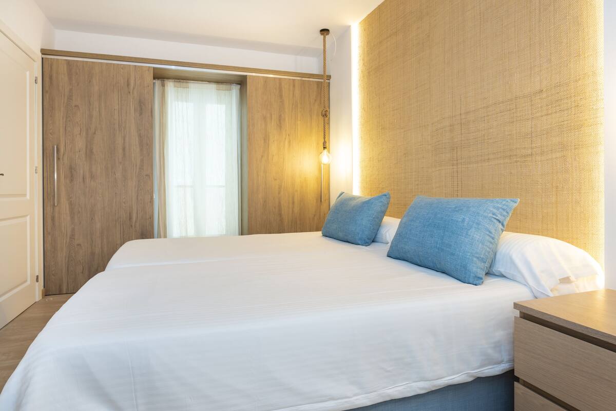 Uma cama de casal com lençóis brancos e duas almofadas azuis, uma cabeceira de madeira, uma mesinha de cabeceira com abajur e um armário com portas de correr.