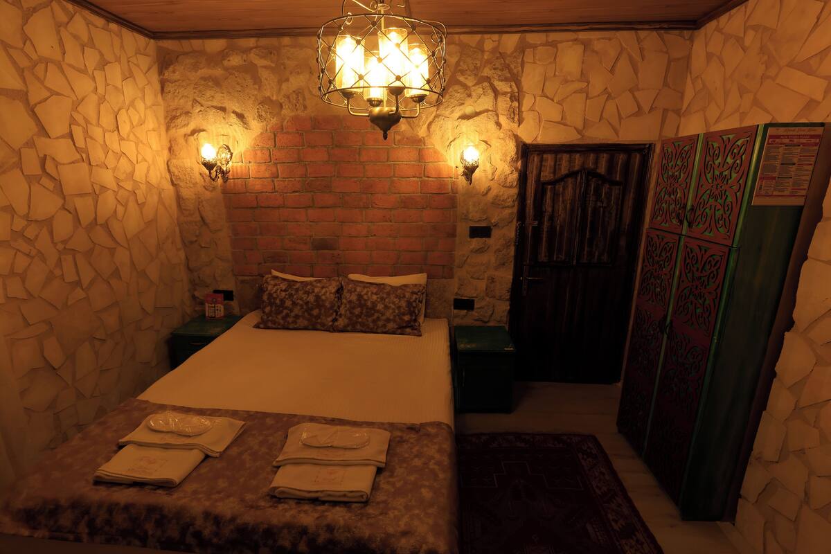 Quarto com parede de pedra, lareira de tijolos, cama com toalhas, mesa de cabeceira verde e porta decorativa.