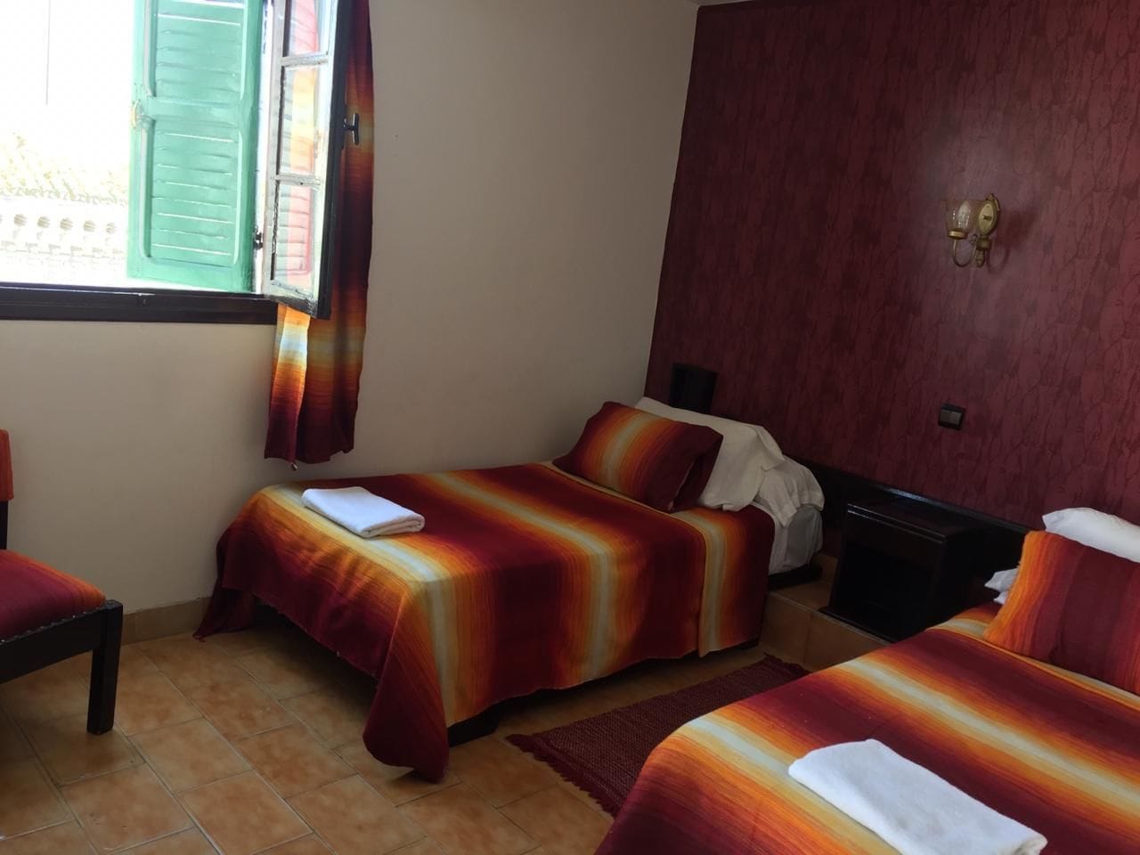 Quarto de hotel com duas camas, janela com persianas verdes, parede vermelha e mesa de cabeceira com abajur.