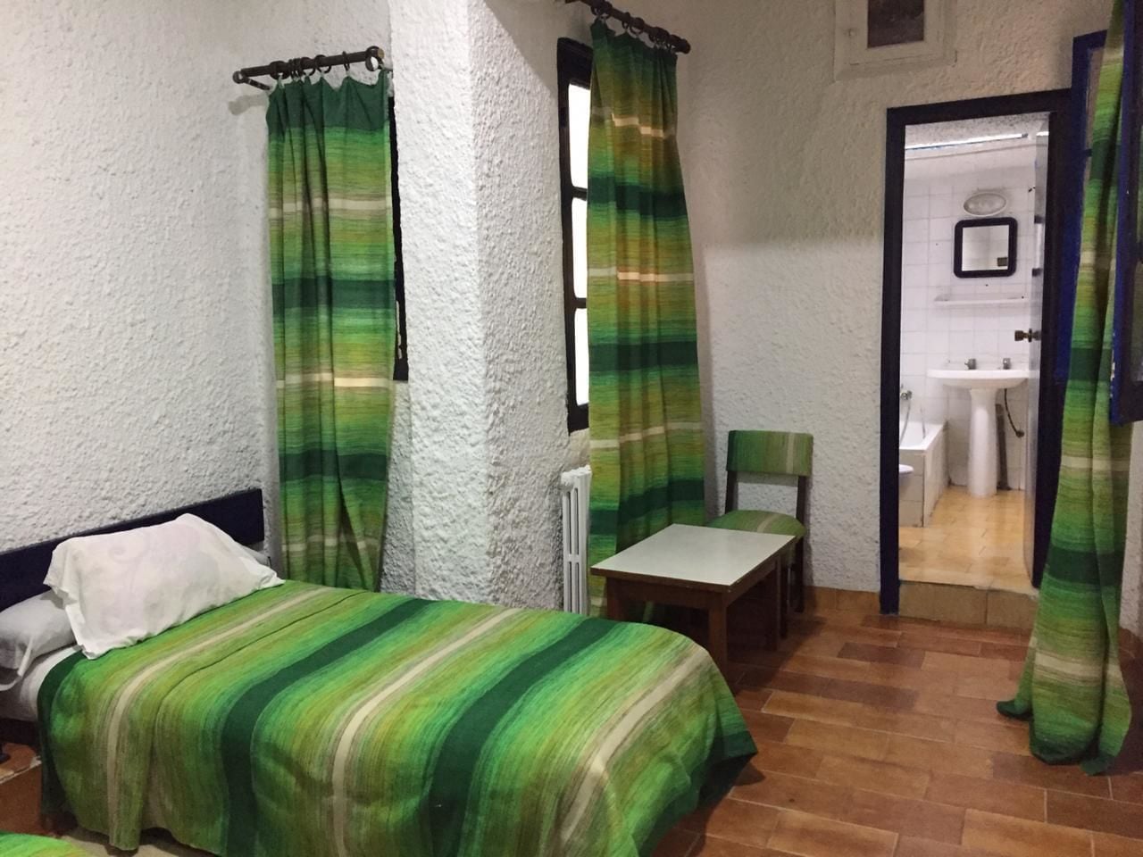Quarto com uma cama, uma mesinha, uma cadeira e banheiro visível por uma porta aberta.