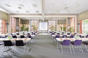 Uma sala de conferências com várias mesas e cadeiras, amplas janelas com cortinas e uma tela de projeção na frente.