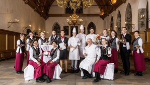 Um grupo de garçons em um ambiente de jantar formal, com um candelabro e janelas em arco.