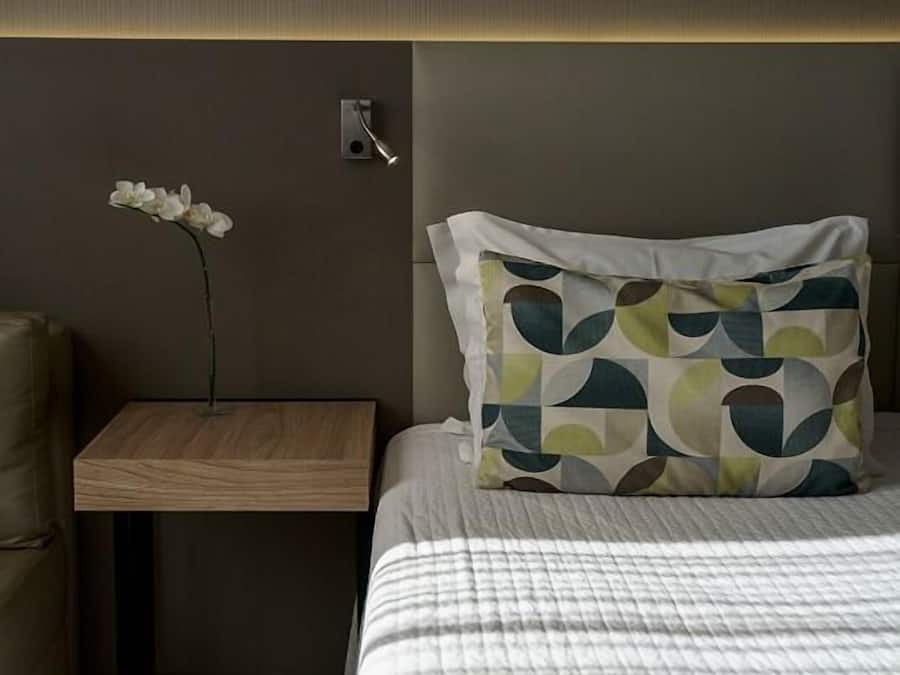 Cama bem arrumada com travesseiro estampado, mesa de cabeceira pequena com um vaso de flores e luminária fixada na parede.
