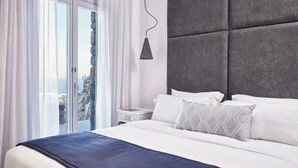 Quarto de hotel moderno com uma cama grande, cabeceira cinza estofada, roupa de cama branca e vista através de uma porta de vidro.