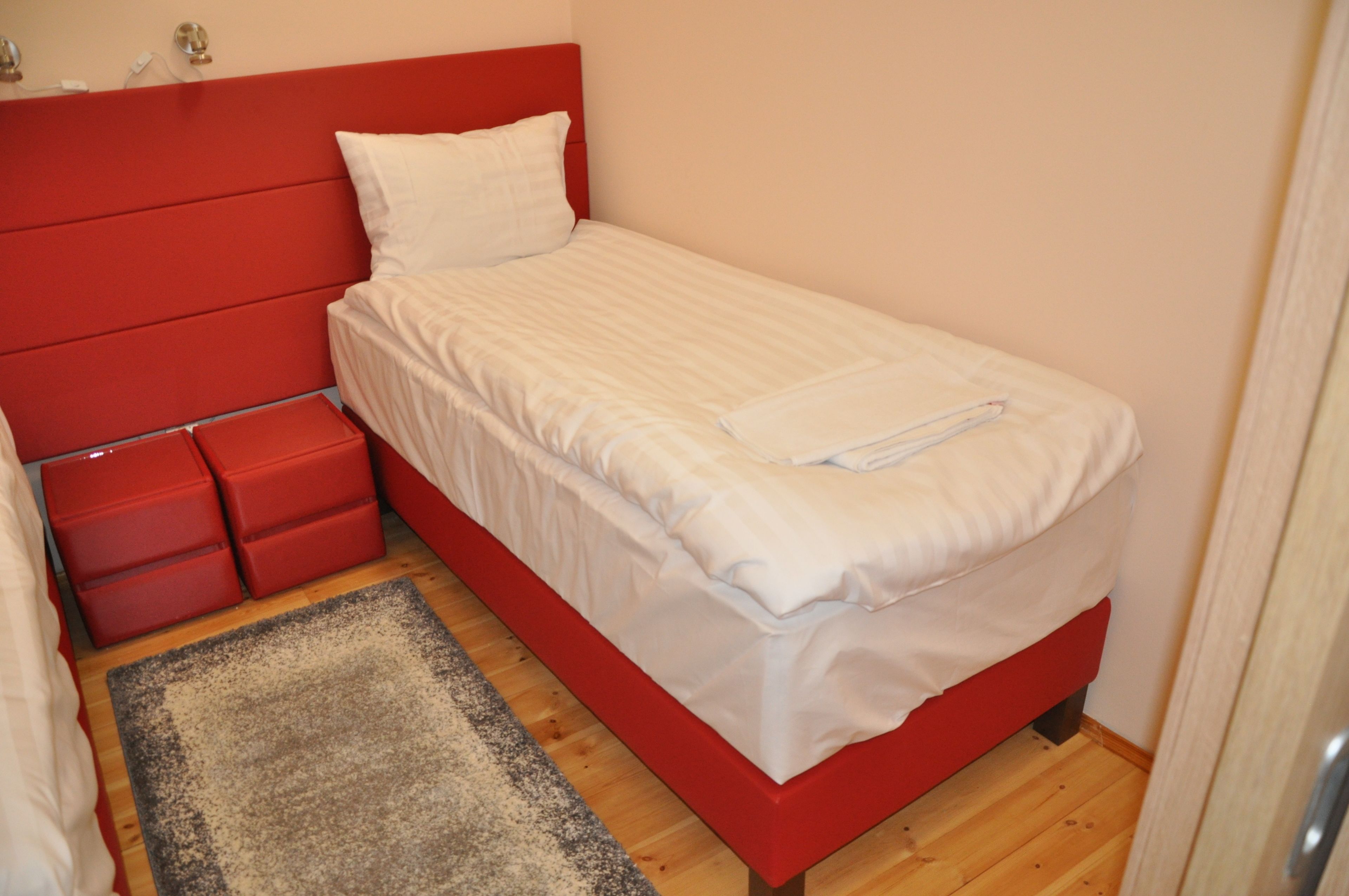 Uma cama pequena com estrutura vermelha e roupa de cama branca em um quarto compacto, com piso de madeira e tapete cinza.