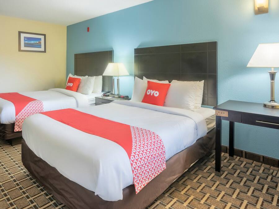 Quarto de hotel com duas camas, cada uma com colcha vermelha e branca, uma mesinha de cabeceira com abajur e uma escrivaninha com luminária.