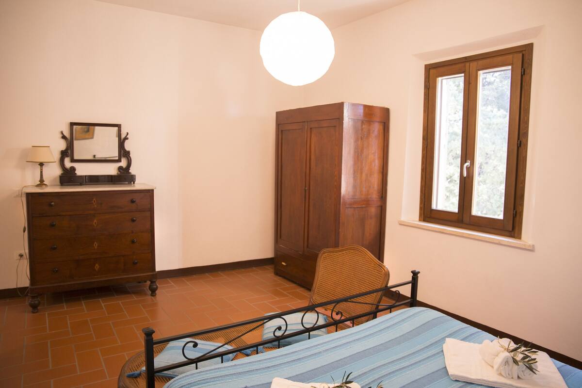 Quarto com cama, guarda-roupa de madeira, cadeira, penteadeira, espelho e janela.