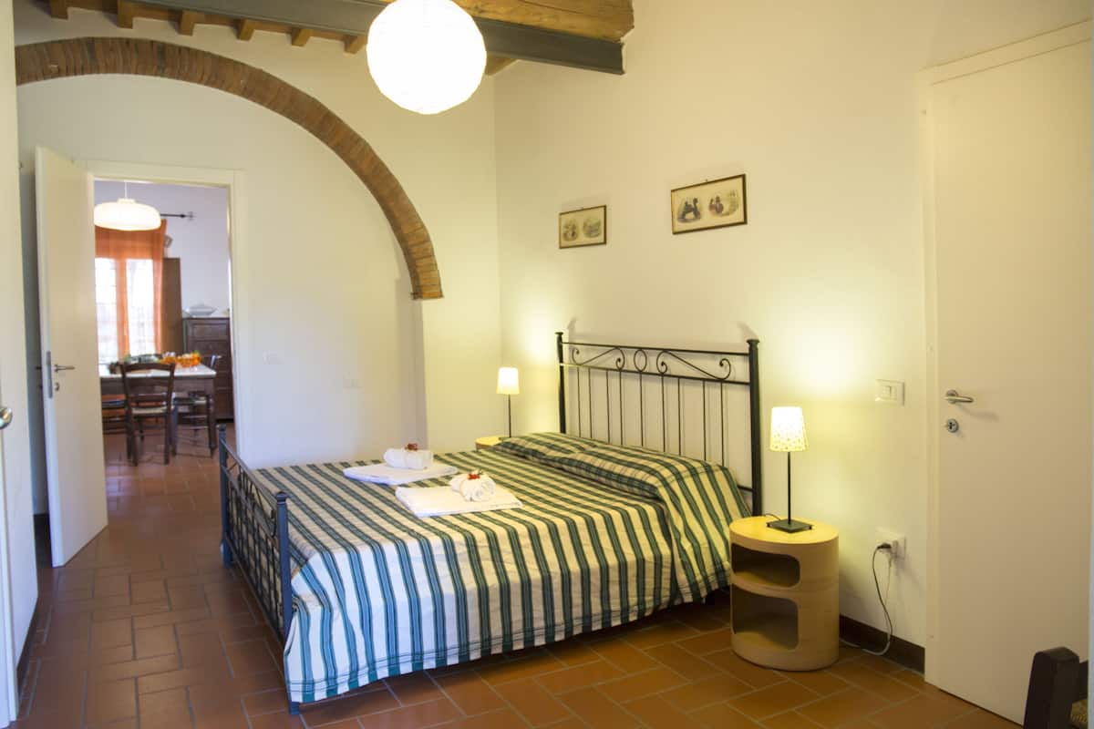 Quarto com cama, luminárias de cabeceira, uma mesinha e uma porta.