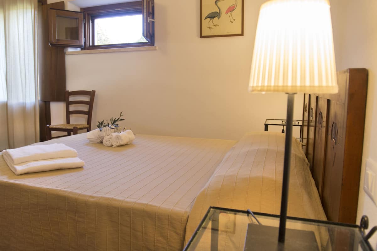 Uma cama bem arrumada com toalhas dobradas, um abajur, uma janela com cortinas, uma cadeira de madeira e um quadro na parede.