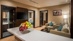 Quarto de hotel moderno com uma televisão de tela plana grande, um sofá confortável, área de refeições com frutas e uma pintura na parede.