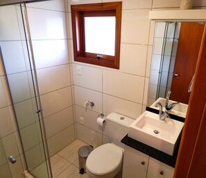 Casa de banho com chuveiro, sanita, lavatório e uma janela sobre o lavatório.