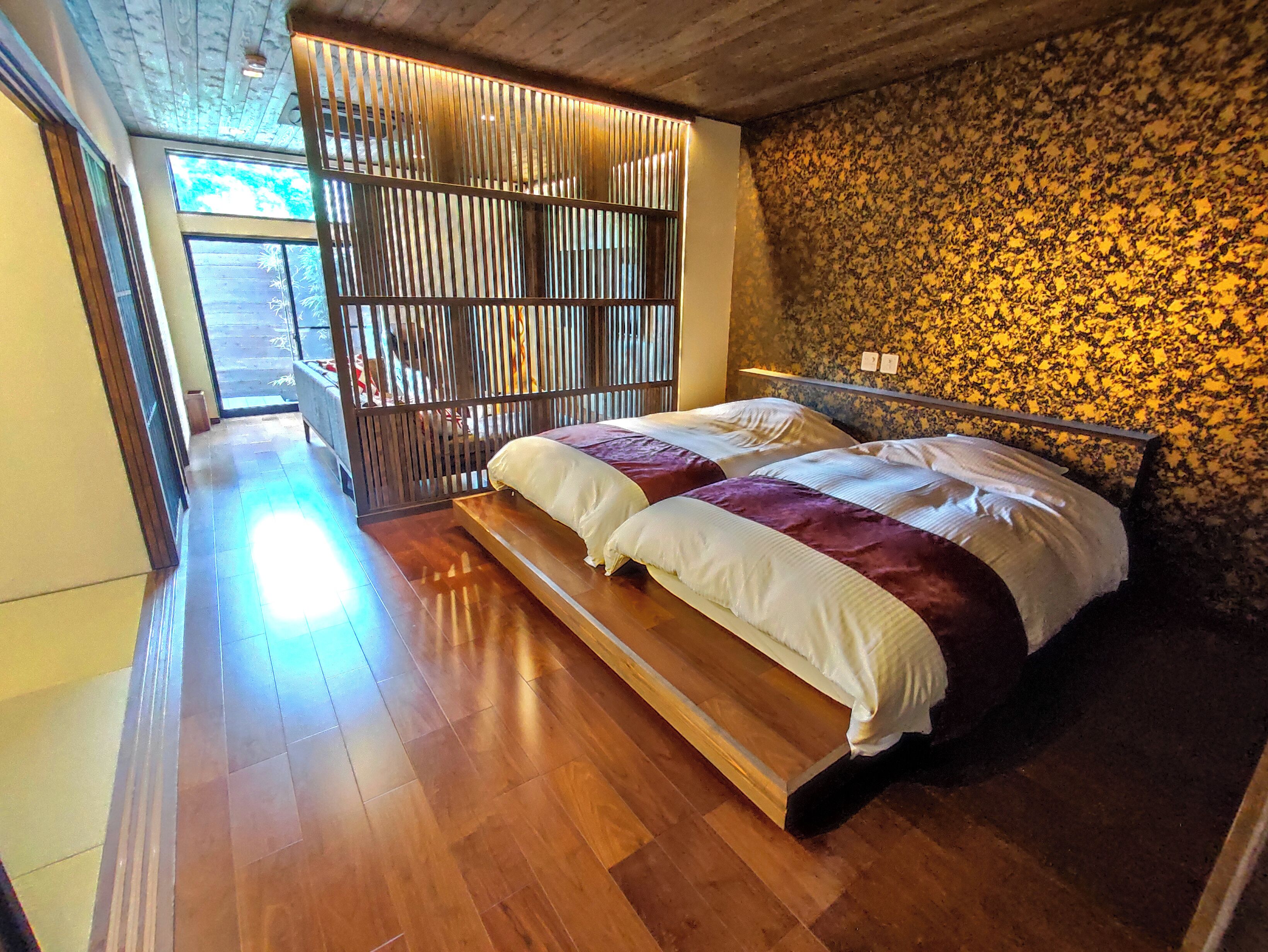 Quarto com piso de madeira, cama com roupa de cama branca e roxa, cabeceira de madeira e janela com persianas.