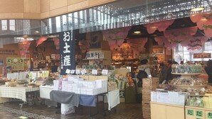Uma banca de mercado com diversos produtos expostos, uma placa com texto em japonês e uma pessoa ao fundo.