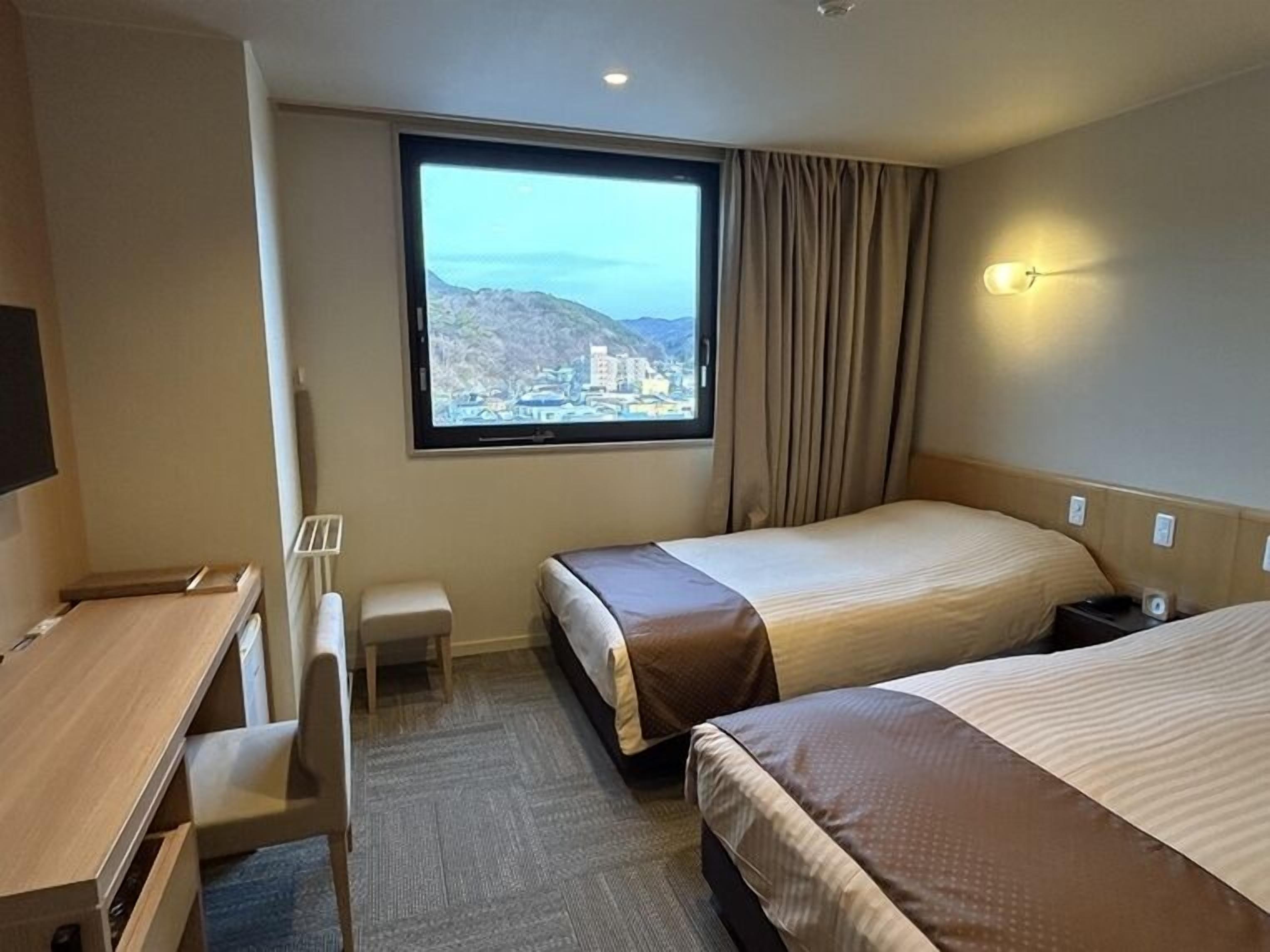 Quarto de hotel com duas camas, uma escrivaninha, uma cadeira, uma janela com vista para a paisagem urbana e uma luminária na parede.