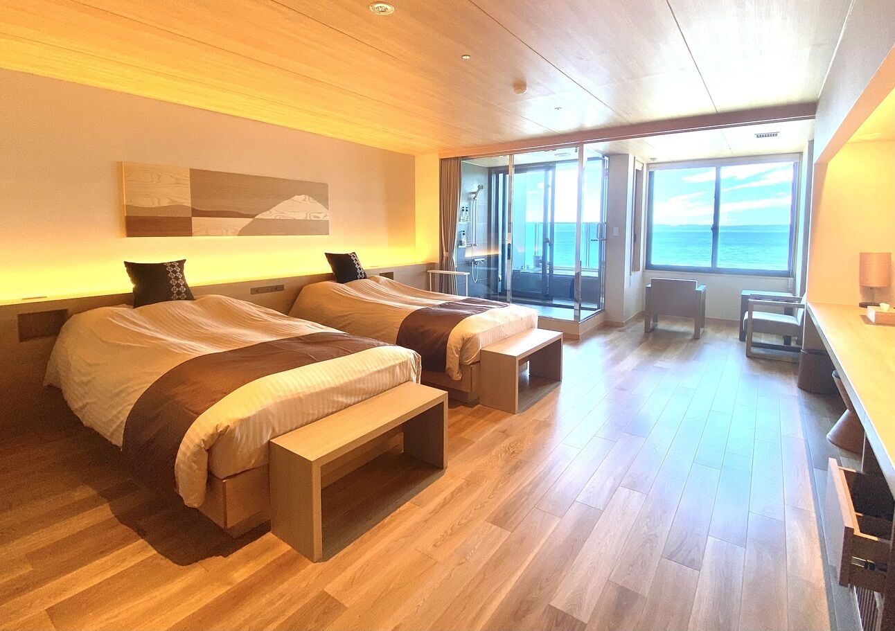 Quarto de hotel moderno com duas camas, uma janela grande e varanda com vista para o mar.