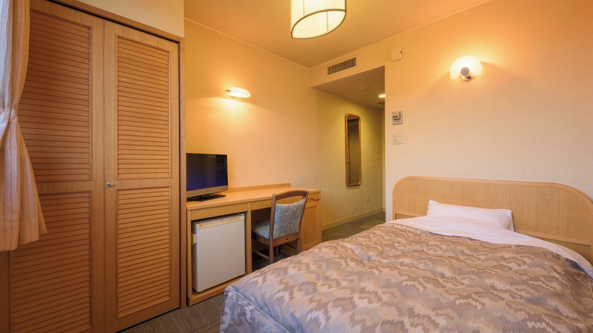 Quarto de hotel com cama, escrivaninha, televisão, cadeira e guarda-roupa.