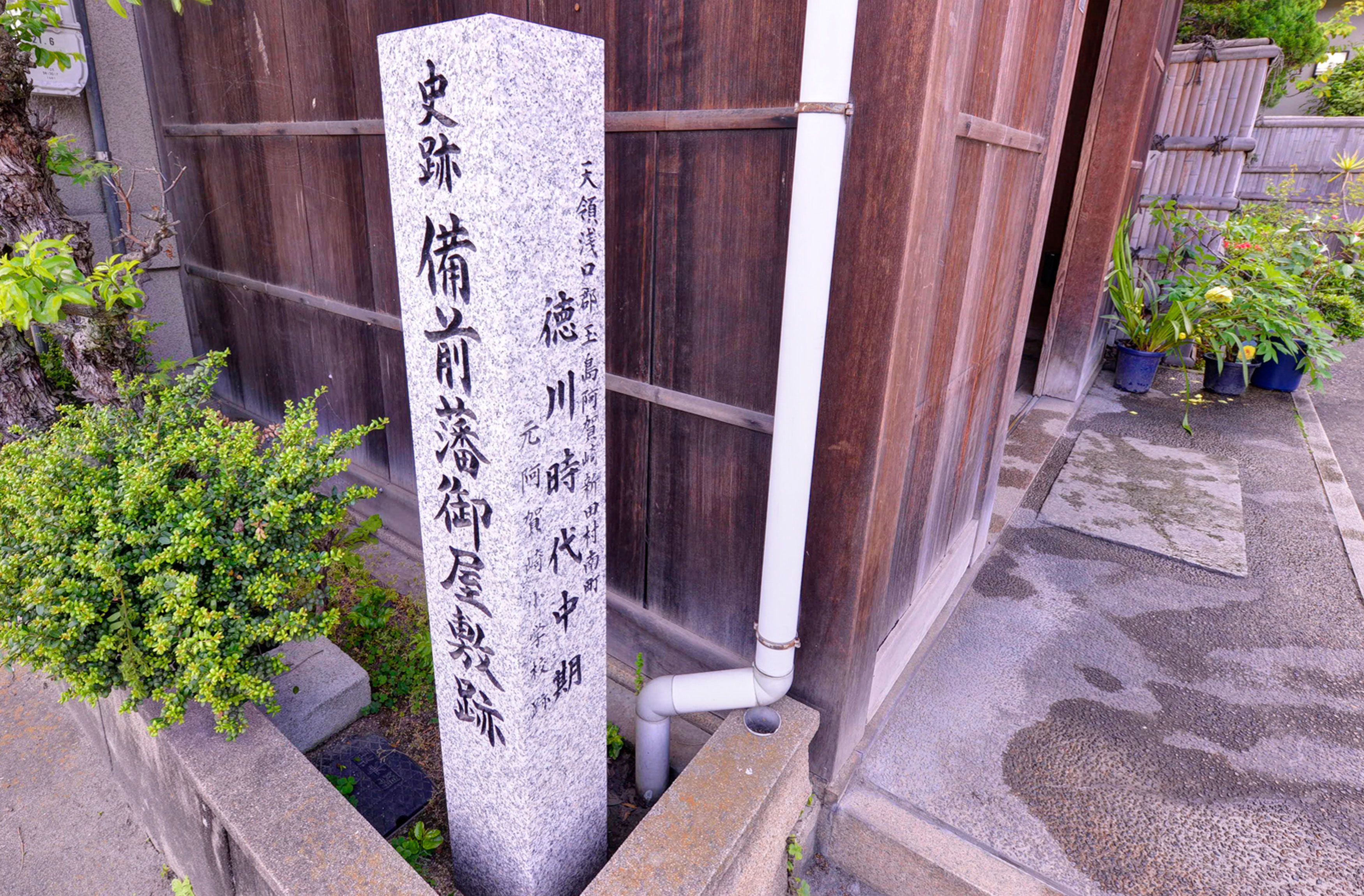 Um monumento de pedra com caracteres japoneses, uma cerca de madeira e plantas em vasos.