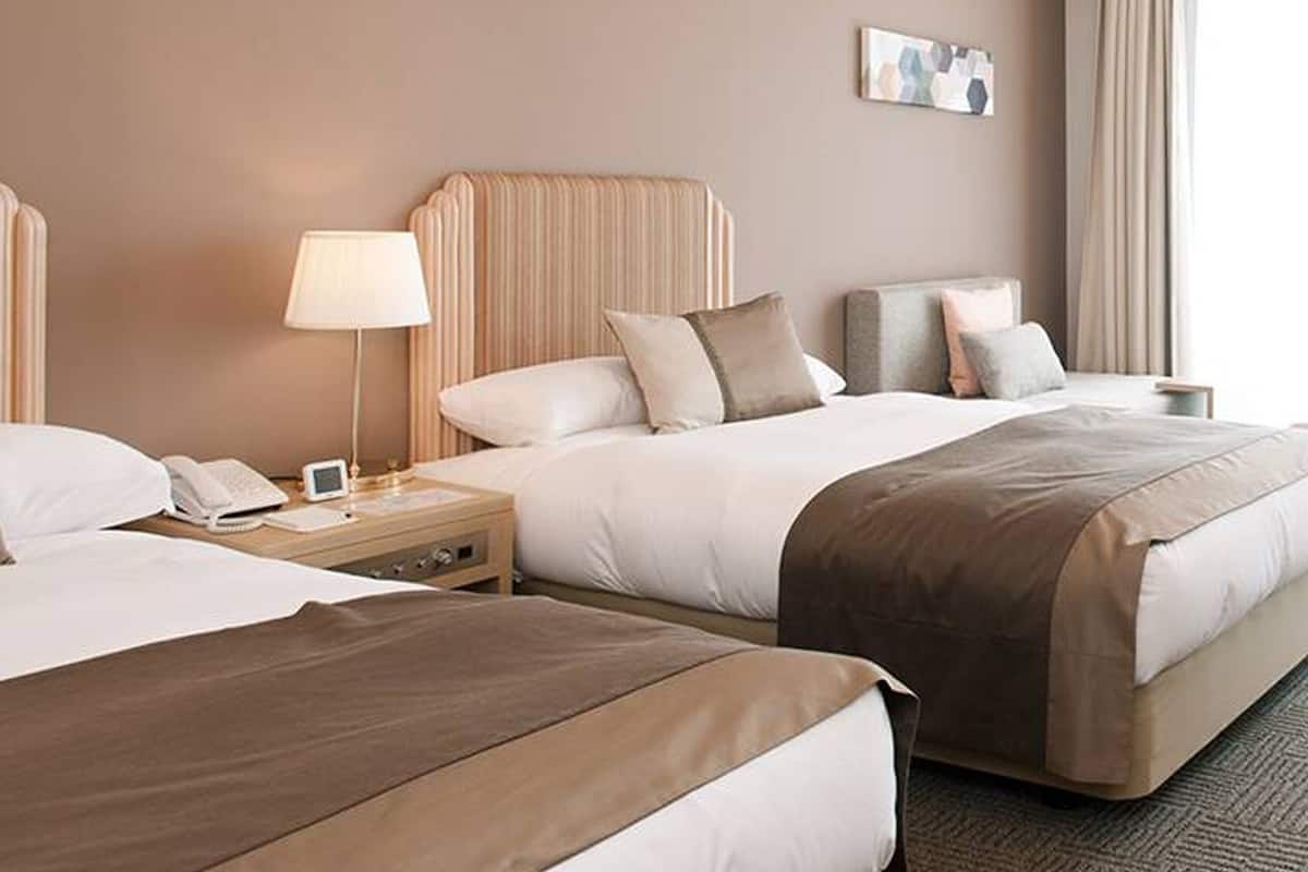Quarto de hotel com duas camas, mesa de cabeceira com abajur, telefone e uma tela pequena.