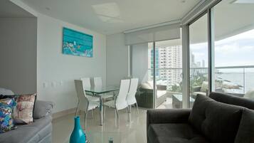 Uma sala de estar moderna com uma mesa de jantar de vidro, cadeiras brancas, um sofá cinza e uma grande janela com vista para a cidade.
