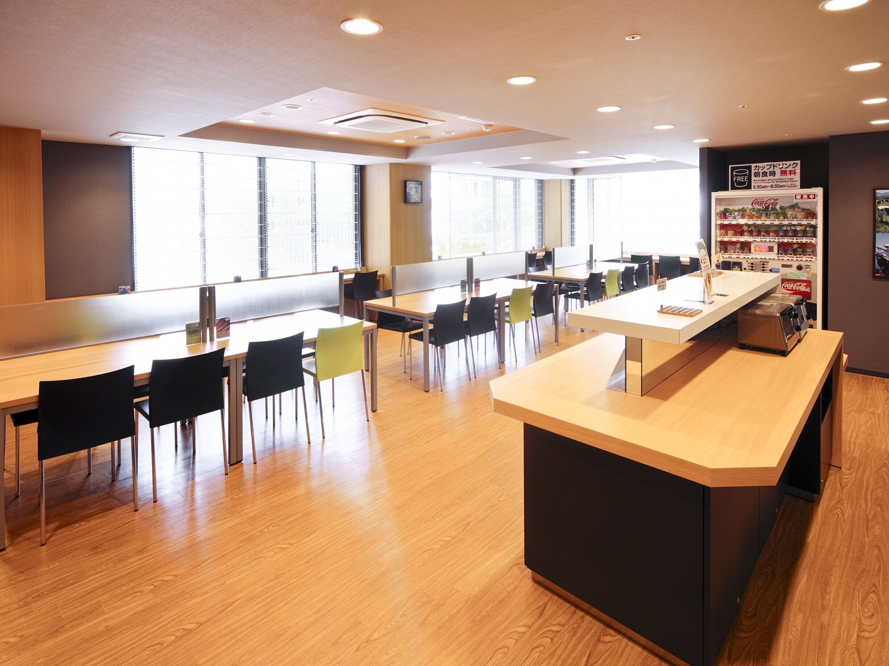 Uma cafeteria moderna com mesas e cadeiras compridas, uma máquina de vendas automática e uma grande janela que proporciona iluminação natural.