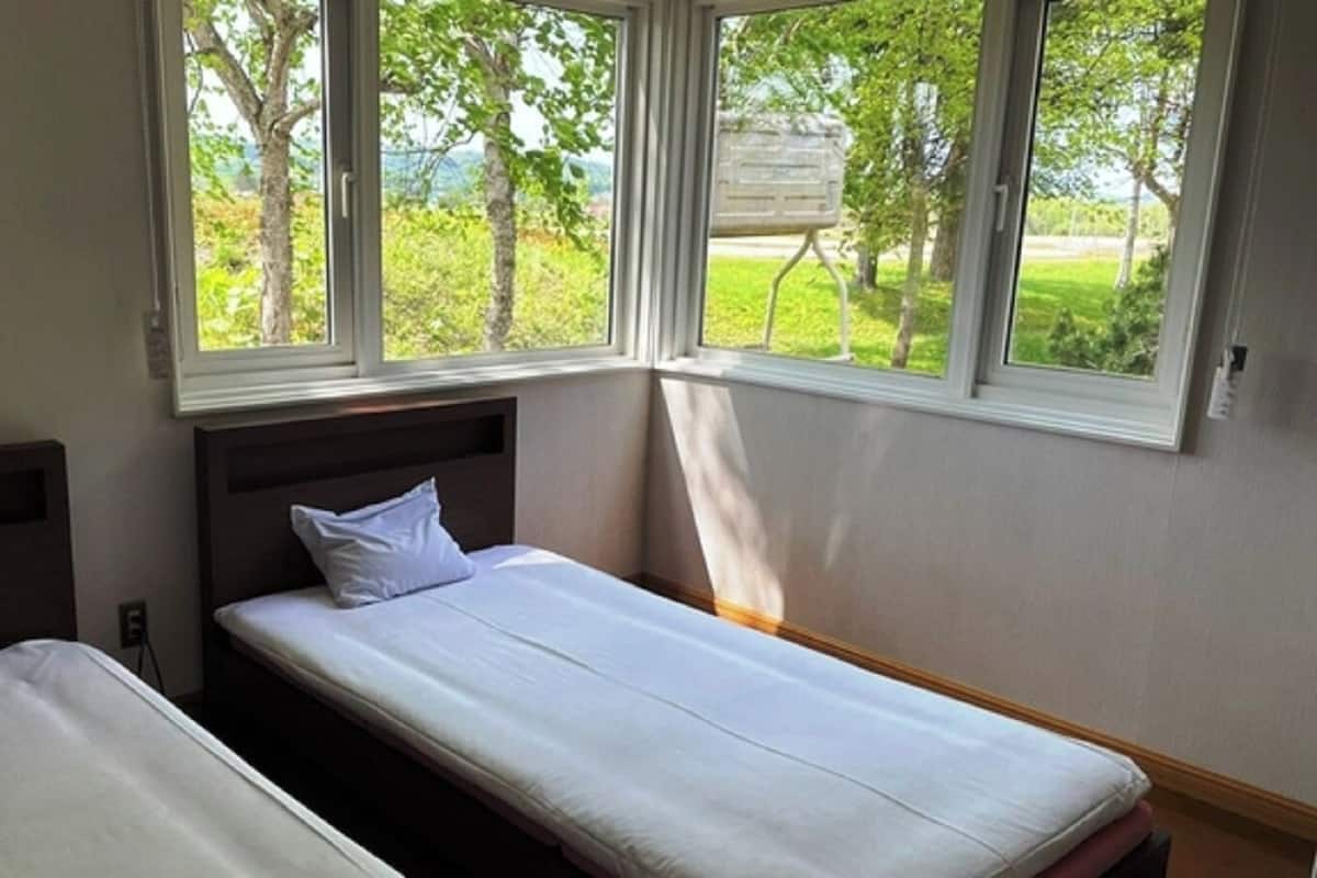Cama de casal com roupa de cama branca e cabeceira de madeira em quarto com grande janela dando vista para árvores e área gramada.