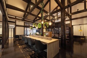 Uma área de bar com um balcão comprido e vários bancos, teto com vigas aparentes e uma estante de livros ao fundo.