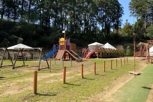 Uma área de recreação infantil com diversos brinquedos, incluindo escorregadores e balanços, cercada por uma grade.
