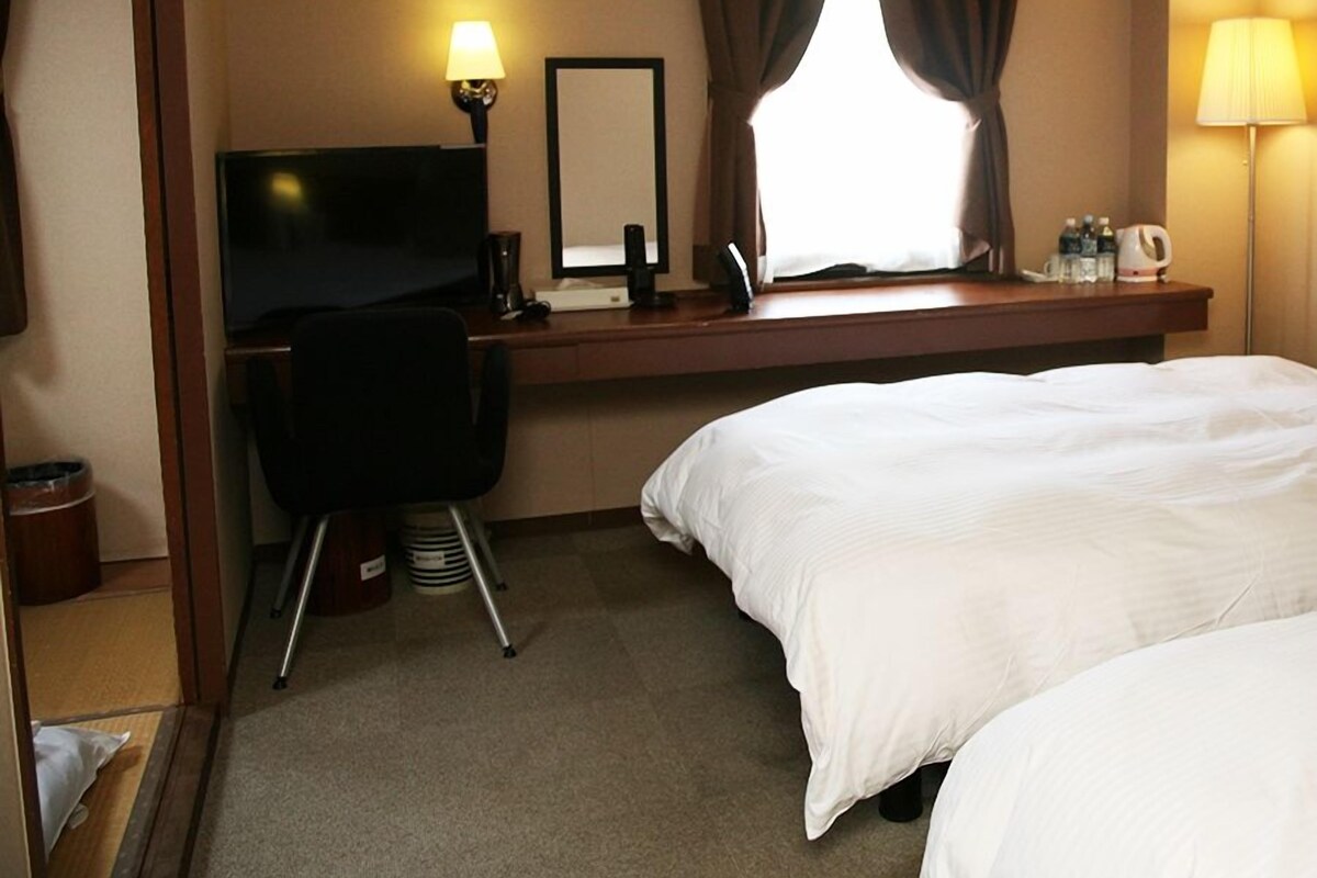 Quarto de hotel com cama, escrivaninha, cadeira, computador, abajur e janela com cortinas.
