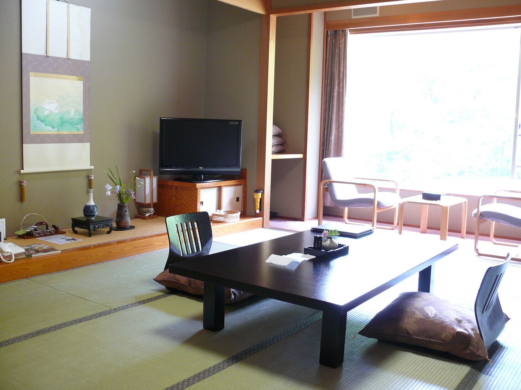 Uma sala de estar tradicional japonesa, com piso de tatame, uma mesa baixa, uma televisão e uma porta deslizante que dá para uma varanda.