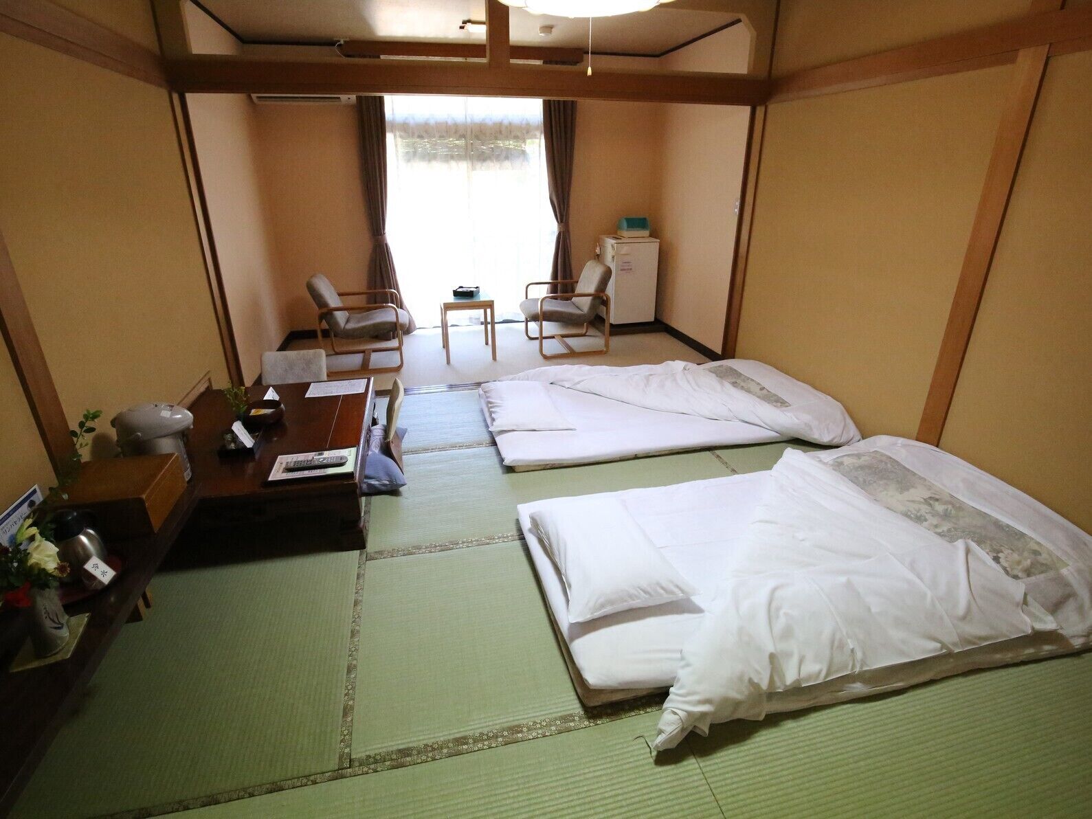 Quarto de hotel com duas camas, uma escrivaninha com cadeira, uma mesinha e uma janela com cortinas.