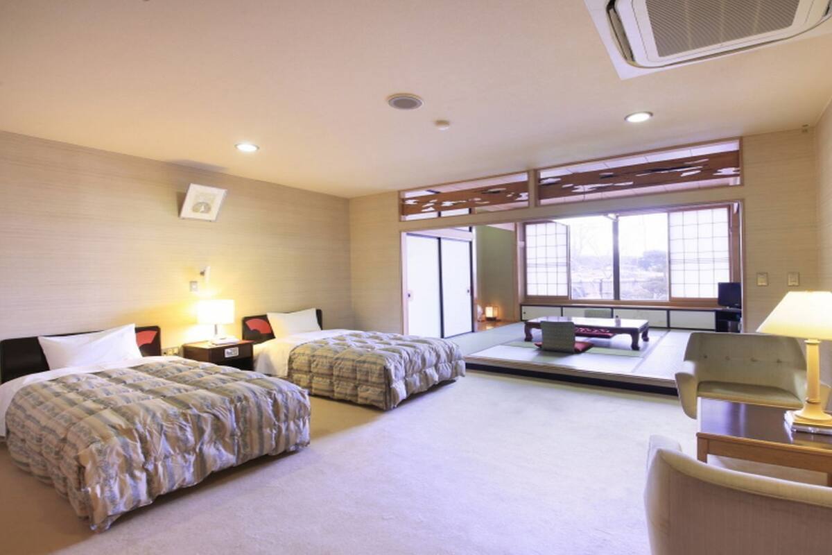 Quarto de hotel com duas camas, uma escrivaninha, uma cadeira, uma janela e uma porta tradicional japonesa deslizante.