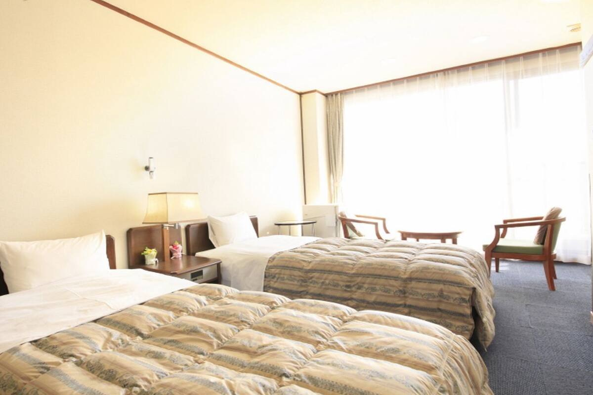 Quarto de hotel com duas camas, uma mesa de cabeceira com abajur, uma cadeira e vista para o exterior pela janela.