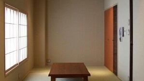 Um quarto com piso de tatame, uma mesa de madeira baixa, uma porta deslizante e uma janela com persianas verticais.