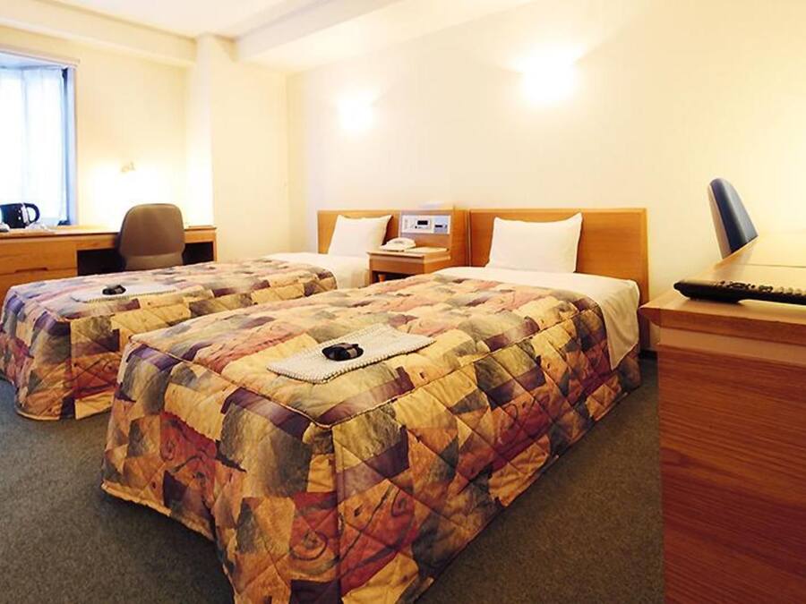 Quarto de hotel com duas camas, uma escrivaninha, uma cadeira, uma TV e uma janela.