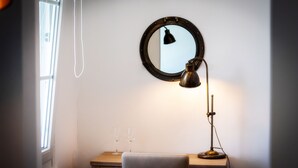 Um quarto pequeno com um espelho redondo, uma escrivaninha com dois copos e uma luminária.