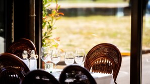 Mesa de jantar posta com cadeiras, copos e garrafas, com vista para uma área gramada pela janela.