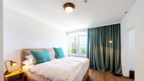 Quarto com cama, mesa de cabeceira, abajur, janela com cortinas e vista para o verde lá fora.