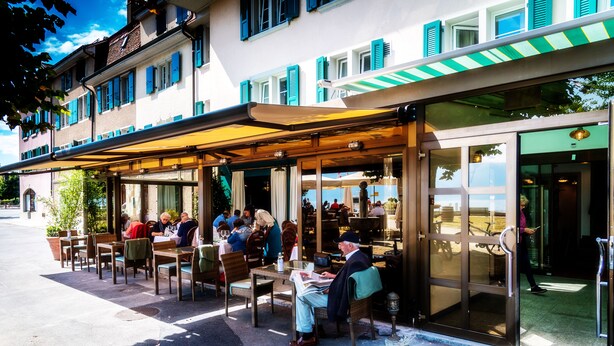 Um café ao ar livre com mesas e cadeiras, uma área de estar coberta e um edifício com janelas e portadas grandes.
