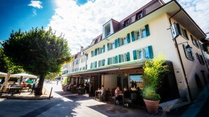 Um café com mesas ao ar livre, uma árvore e um edifício com várias janelas e varanda.