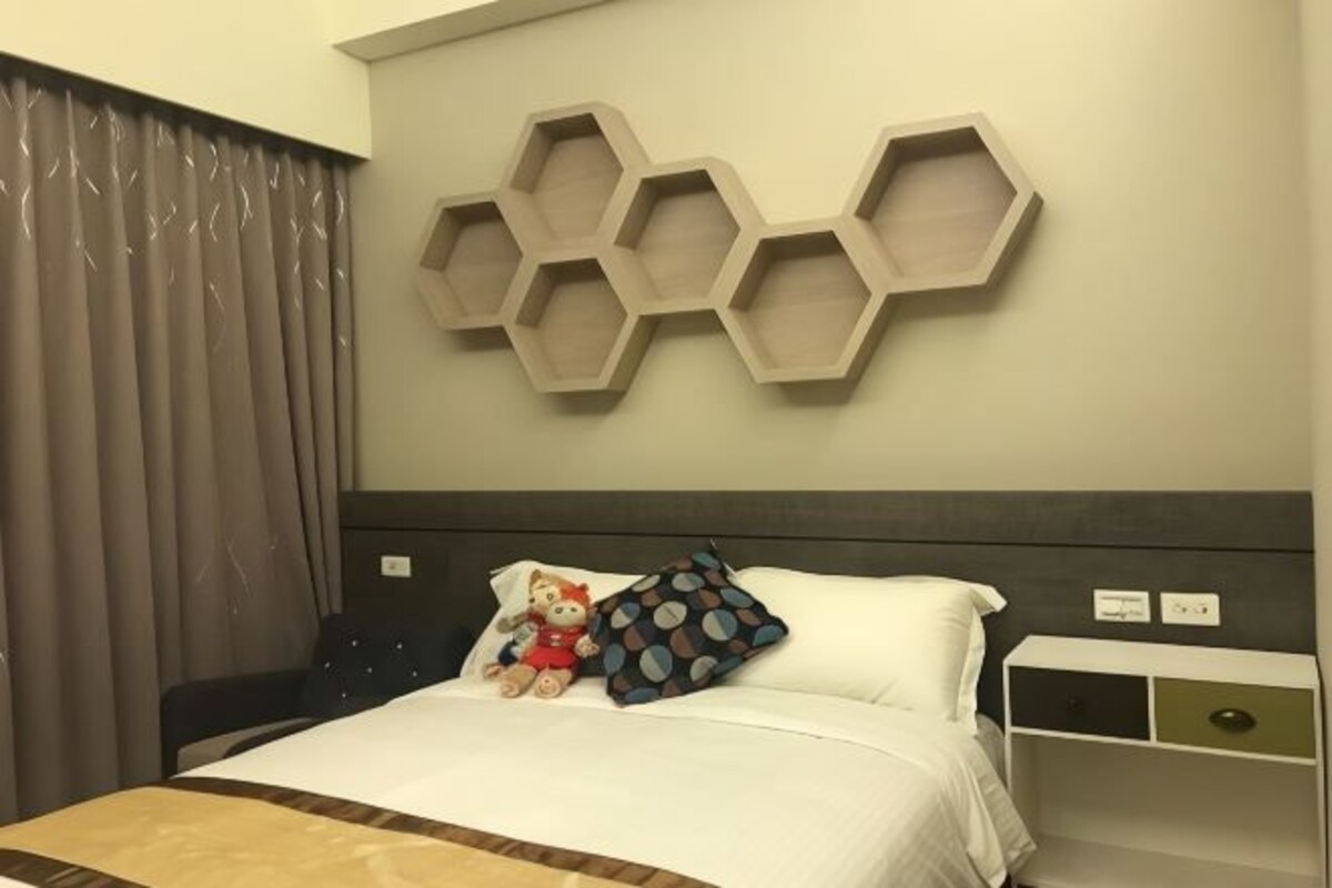 Quarto de hotel com cama, cabeceira, mesa de cabeceira e prateleira hexagonal fixada na parede.