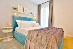 Quarto com cabeceira azul, roupa de cama branca, mesa de cabeceira com abajur e janela com cortinas.