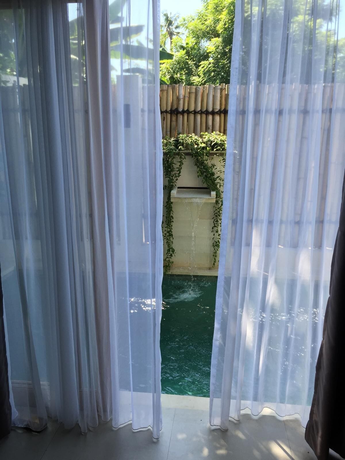 Visão através de cortinas finas para uma área de piscina com cerca de bambu e vegetação.
