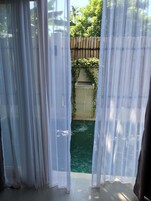 Visão através de cortinas finas para uma área de piscina com cerca de bambu e vegetação.