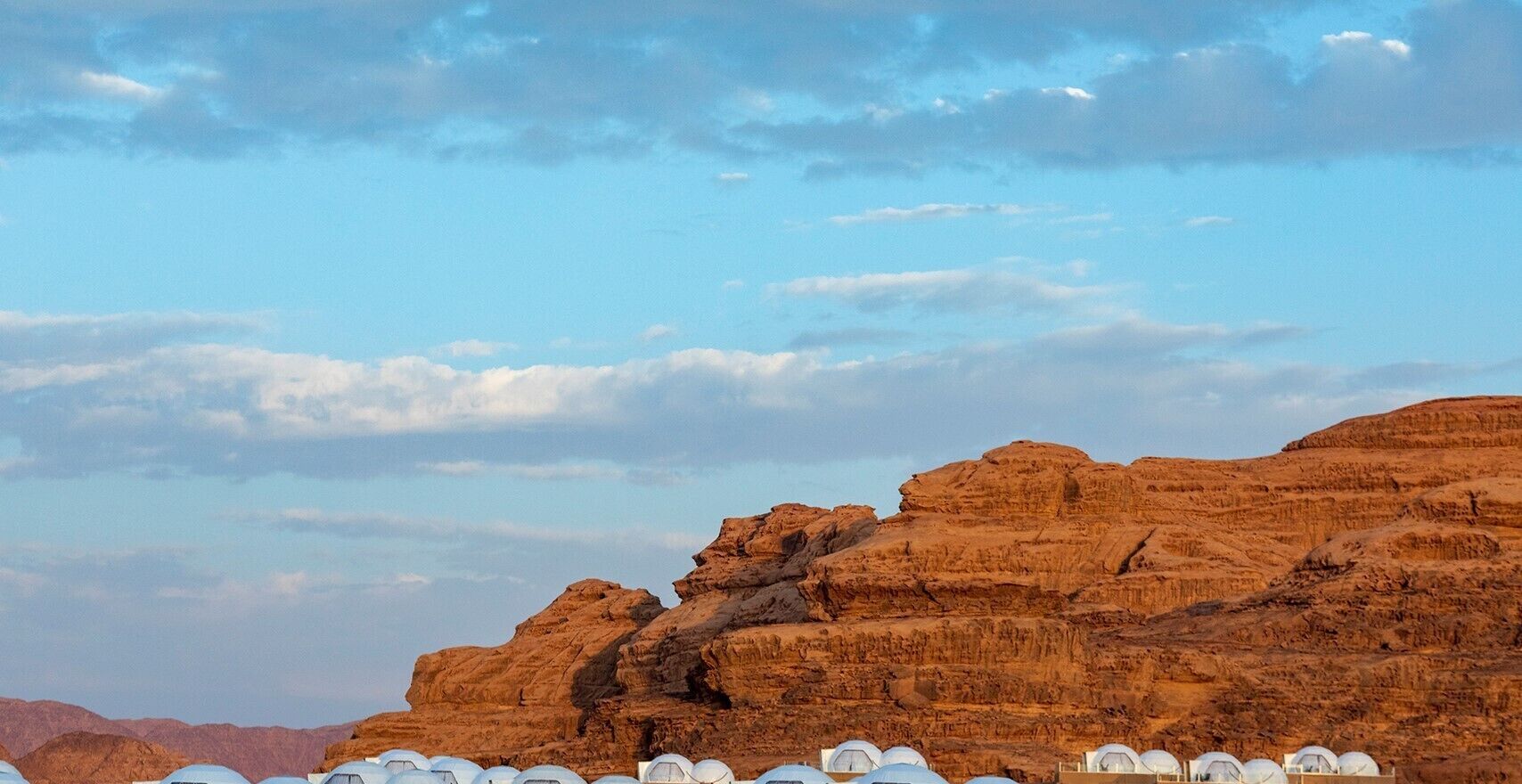 Paisagem desértica com uma colina rochosa destacada e várias estruturas em forma de cúpula na base.