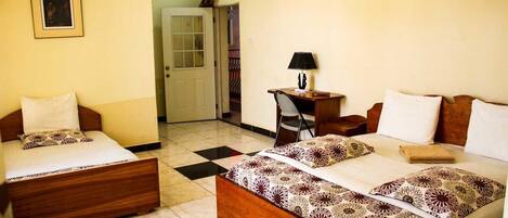 Quarto de hotel com duas camas de solteiro, uma mesa com cadeira, uma luminária e uma porta que dá para uma varanda.