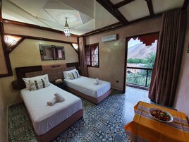 Quarto de hotel com duas camas, um ventilador de teto, uma janela com vista para montanhas e uma mesa com frutas.