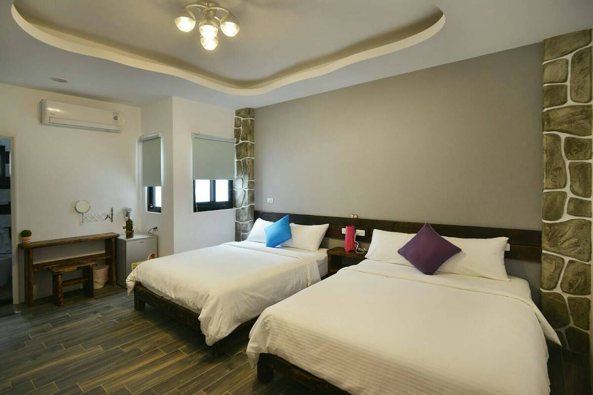 Quarto de hotel com duas camas, uma pequena escrivaninha, um espelho e uma parede de destaque em pedra.