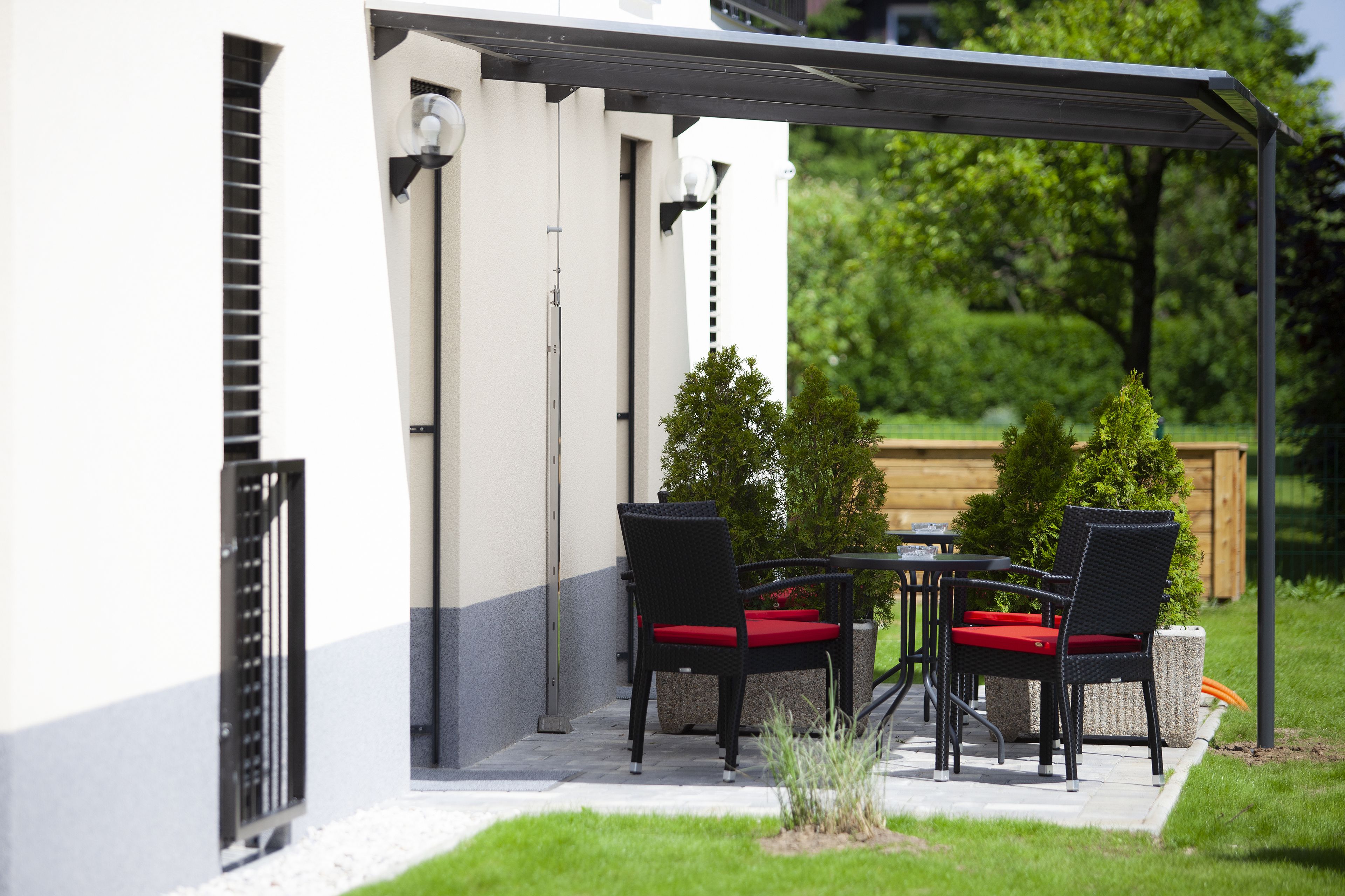 Um pátio externo com mesa e cadeiras, área coberta e um pequeno jardim com cerca de madeira ao fundo.