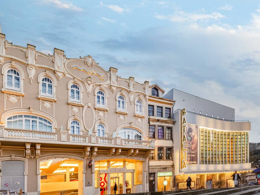 Um edifício histórico com arquitetura ornamentada, uma extensão moderna em vidro e a placa de um hotel.