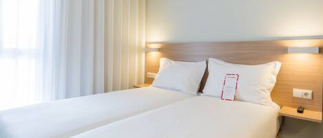 Uma cama de casal com lençóis brancos, cabeceira de madeira, mesas de cabeceira com abajures e uma grande janela com cortinas translúcidas.